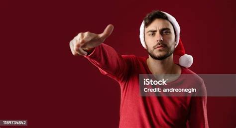 빨간 스웨터와 산타 모자를 입은 젊은 잘생긴 백인 남자가 스튜디오에서 빨간색 배경에 서서 카메라에 손가락을 보여줍니다 12월에