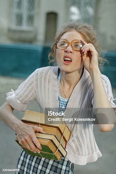 커다란 안경을 쓴 괴짜 젊은 여성은 무거운 책을 들느라 고군분투하고 햇빛 아래 야외에 서 있으면서 지쳐서 하품하거나 헐떡입니다 20 24세에 대한 스톡 사진 및 기타 이미지