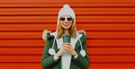 빨간 배경에 폼 폼이 달린 흰색 모자를 쓰고 커피 컵을 들고 미소짓는 젊은 여성의 겨울 초상화 20 29세에 대한 스톡 사진 및