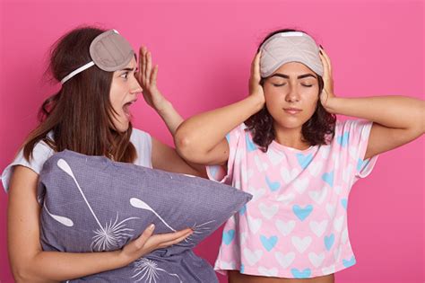충격으로 입을 크게 벌리고 베개를 손에 들고 손으로 이마를 만지고 친구를 바라보며 비명을 지르는 젊은 여성의 프로필 검은 머리 아가씨는 닫힌 눈과 귀로 포즈를 취합니다