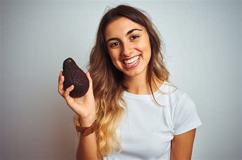 행복한 얼굴 서서 치아를 보여주는 자신감 미소와 함께 회색 고립 된 배경을 통해 아보카도를 먹는 젊은 아름다운 여자 건강한 생활방식에 대한 스톡 사진 및 기타 이미지