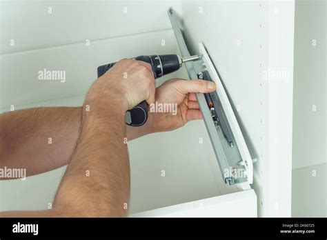 Male Hands Hold Cordless Electric Screwdriver Screw To Fix Rail Assembling Drawer Of White
