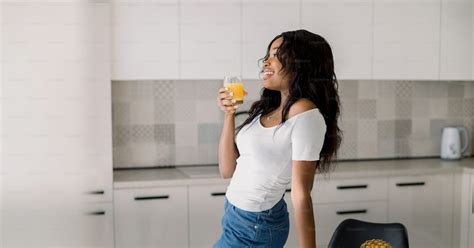 젊은 아름다운 아프리카계 미국인 여자는 오렌지 주스가 든 유리를 들고 창문을 바라보며 집에서 가벼운 현대적인 부엌에서 포즈를 취하고 있습니다 건강한 라이프스타일 컨셉