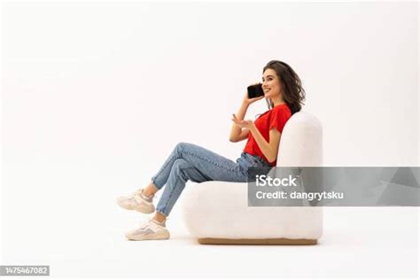 웃고 있는 젊은 여자가 전화로 대화를 나누고 하얀 스튜디오 벽에 아늑한 의자에 앉아 있다 스마트폰으로 고객이나 친구와 이야기하는 아름다운 백인 여성 말하기에 대한 스톡 사진