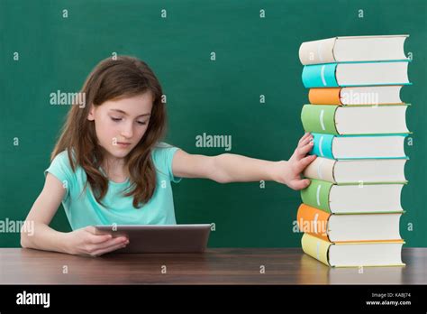 Girl Using Digital Tablet And Avoiding Stack Of Books At Desk In