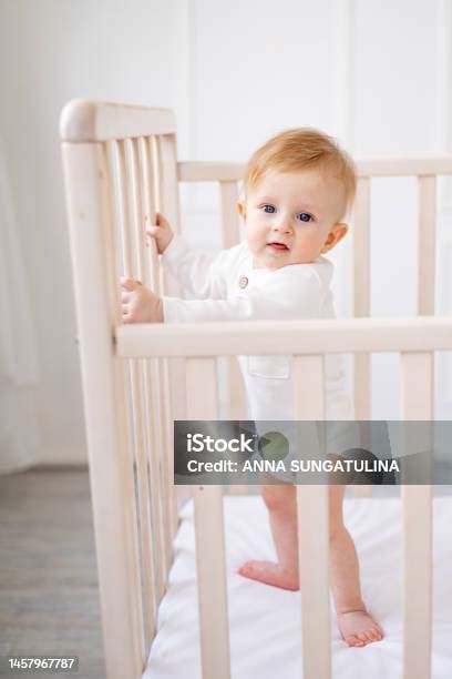 웃는 아기 6 개월 된 금발 소년은 흰색 면화 바디 수트 어린이 용품의 개념으로 밝은 침실의 유아용 침대에 서 있습니다 6 11