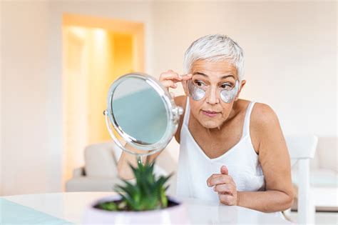 붓기 주름 다크 서클을 위한 아이 마스크 아래 눈 패치 개념 흰색 셔츠를 입은 수석 회색 머리 의 여성 거울을 보면서 실버 눈