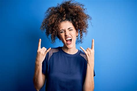 곱슬 머리와 피어싱 캐주얼 블루 티셔츠를 입고 젊은 아름다운 여자는 손을 위로 바위 기호를하고 미친 표정으로 외칩니다 음악 스타