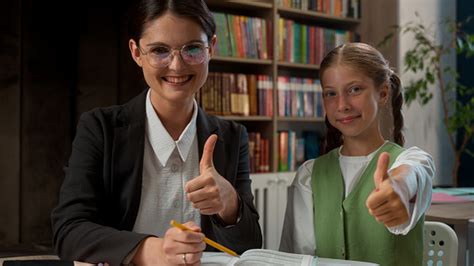 교육자 교사 백인 여성 여성 교수 교사 도움 숙제 과제 설명 교육 학습 어린 소녀 학교 아이 여학생 여학생 보고 카메라 쇼 엄지 손가락 위로 도서관 2명에 대한 스톡 사진