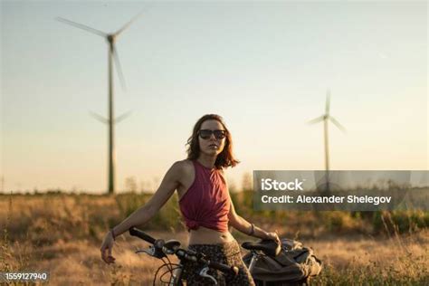 해질녘 야외에서 산악 자전거를 탄 아름다운 여성의 여성 초상화 아름다운 사람들과 함께하는 라이프스타일 사진 선글라스를 쓴 사람 풍력 발전 단지의 터빈 녹색 에너지 기술로