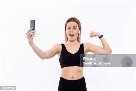 검은 스포츠웨어에 젊은 스포티 한 금발 의 여자 흰색 배경 위에 서 운동 후 이두근을 보여주는 스마트 폰과 셀카를 만들기 무선 헤드폰으로 운동 중 음악을 듣는 모델 달리기에