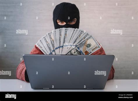 A Male Thief Holds Dollars In His Hand Using A Laptop Computer For