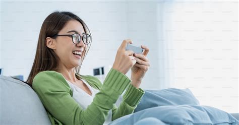 안경을 쓴 아시아 여성은 집에서 스마트폰으로 모바일 게임을 한다 매력적인 캐주얼 소녀는 행복하고 긴장을 풀고 소파에 앉아 전화로 화면을 만지는 재미를 가지고 집에서 승리를