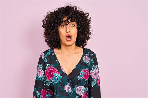 고립 된 분홍색 배경에 꽃 드레스를 입고 곱슬 머리를 가진 젊은 아랍 여성은 놀라움의 표정 두려움과 흥분 된 얼굴로 충격을 받았습니다 감정에 대한 스톡 사진 및 기타 이미지