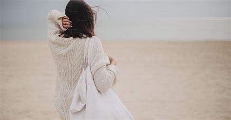 바람이 부는 머리와 토트백을 들고 모래 해변에서 바다로 걸어가는 아름다운 세련된 여성 평온한 순간 니트 스웨터를 입은 세련된 젊은 여성이 휴가를 즐기고 해안에서 휴식을