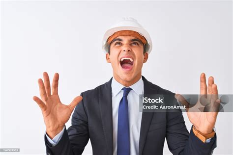 고립된 흰색 배경 위에 양복과 헬멧을 쓰고 있는 젊은 잘생긴 건축가 남자는 공격적인 표정과 팔을 들고 미친 소리와 고함을 질렀다 좌절 개념입니다 감정에 대한 스톡 사진 및