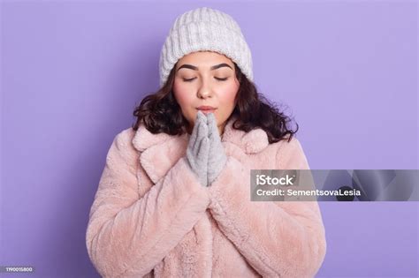 겨울에 손을 잡고 따뜻한 옷 인조 모피 코트 장갑 모자를 쓰고 라일락 배경으로 고립 된 포즈를 취한 아름다운 젊은 여성이 눈을 감고 서 있습니다 겨울과 추운 날씨 개념입니다