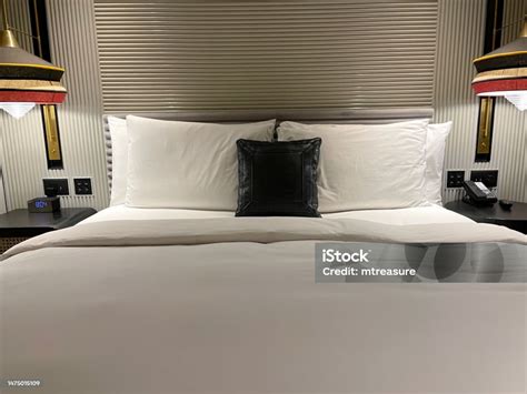 질감이 있는 더블 침대 지휘봉 헤드보드 쌓인 흰색 베개 접힌 이불 침대 협탁의 디지털 알람 시계 조명이 켜진 교수형 펜던트 램프
