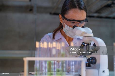 마스크와 장갑을 낀 과학자가 실험실에서 비커에 담긴 녹색 액체를 현미경으로 검사하고 있습니다 실험실 설정은 연구 과학 및 실험에 중점을 둔 밝은 조명입니다 Stem 주제에