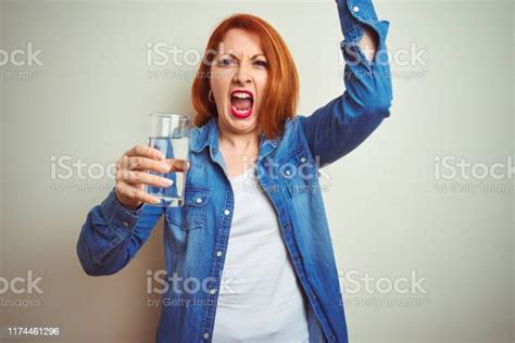 흰색 고립 된 배경을 통해 물 잔을 마시는 젊은 아름다운 빨간 머리 여자는 짜증과 분노와 함께 고함 미친 고함 제기 손으로 소리