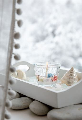 수제 수중 조개 투명 젤 왁스 캔들 투명 컵 안에 집에서 창틀에 바다 유리와 조개껍데기가 있는 흰색 나무 쟁반에 아름다운 촛불