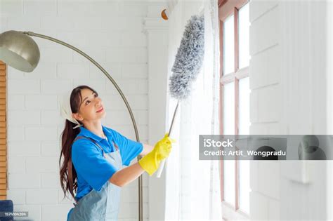 아시아의 젊은 전문 청소 서비스 여성 노동자가 집에서 일하고 있다 소녀는 회색 깃털 먼지떨이로 커튼과 창문을 청소합니다 전문적인 청소부와 집안일 서비스 개념 가사 집안일에