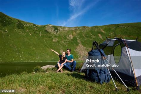 여름 날에 산에 호수에 대 한 캠핑 근처 휴식 하는 젊은 쌍 남자와 여자는 텐트 배낭 트레킹 스틱 근처에 앉아 여자 그녀의 손을 멀리을 보여줍니다 2명에 대한 스톡 사진 및