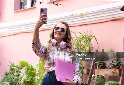 목에 실크 스카프를 두르고 멋진 배경에 포즈를 취하고 벤치에 앉아 녹색 덤불과 보라색 꽃으로 둘러싸여 셀카를 찍는 즐거운 긍정적인 세련된 여성의 야외 사진 60 64세에