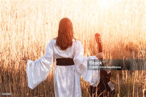 흰색 중세 드레스와 어쿠스틱 기타를 들고 일몰 조명 필드를 통해 걷는 벨트에 빨간 머리를 가진 날씬한 젊은 여성의 다시 보기 렌즈 플레어 가을에 대한 스톡 사진 및 기타