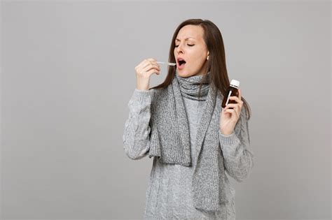 회색 스웨터에 아픈 젊은 여자 스카프는 회색 벽 배경에 고립 된 숟가락에 액체 약을 붓는 다 건강 한 라이프 스타일 아픈 질병 치료 추운 계절 개념입니다 복사 공간을 모의