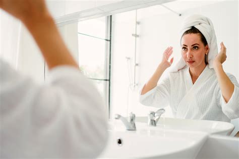 하얀 목욕 가운을 입고 머리에 수건을 쓴 여자가 아침 일과를 하는 동안 욕실 거울 앞에 서 있습니다 밝고 미니멀한 홈 인테리어에서 자기 관리 웰빙 및 위생에 중점을 둡니다