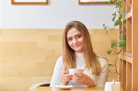 스트레이트 가벼운 머리를 가진 사랑 스러운 젊은 여성 모델의 이미지는 음료의 찻잔을 보유 하 고 자유 시간을가지고 커피 숍에서 파트너와 함께 진정 분위기를 즐긴다 커피를