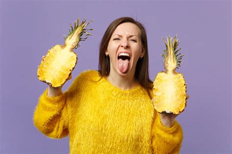 보라색 파스텔 배경에 고립 된 신선한 익은 파인애플 과일의 두 Halfs를 들고 혀를 보여주는 모피 스웨터에 미친 젊은 여자 사람들은 생생한 라이프 스타일 휴식 휴가