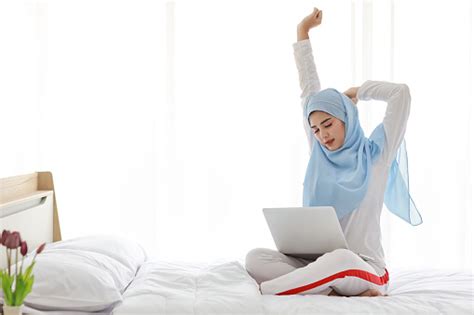 침실에서 침대에 앉아 컴퓨터를 사용하는 젊은 아시아 여성 잠자리에 들기 전에 일을 마치고 팔을 뻗고 있는 파란색 히잡을 입은 무슬림 잠옷을 입은 아름다운 소녀 라이프스타일과