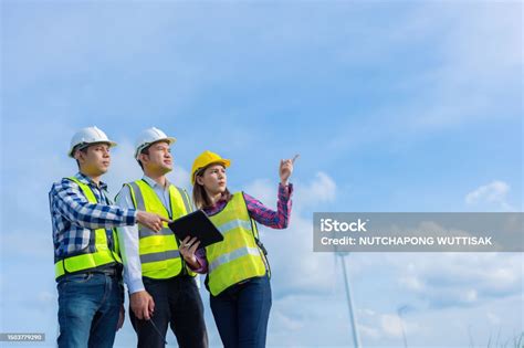 3명의 남녀 엔지니어로 구성된 팀이 희망을 품고 앞을 바라보며 서 있었습니다 청정 에너지 작업을 위해 노트북을 들고 헬멧과 조끼를 착용하십시오 배경에 풍력 터빈이 있습니다