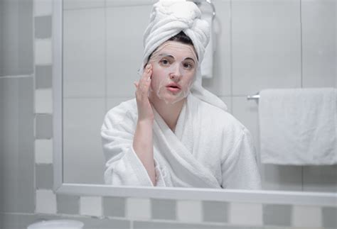 흰색 목욕 가운을 입고 머리에 수건을 두른 젊은 백인 여성이 얼굴에서 보습 시트 마스크를 제거하고 욕실에서 뷰티 트리트먼트를