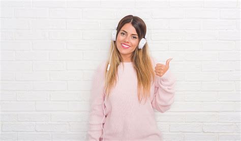 음악을 듣고 춤을 가리키는 및 행복 한 얼굴은 웃 고 옆까지 엄지를 보여주는 헤드폰 착용 백색 벽돌 벽을 통해 젊은 성인 여자 가리키기에 대한 스톡 사진 및 기타 이미지