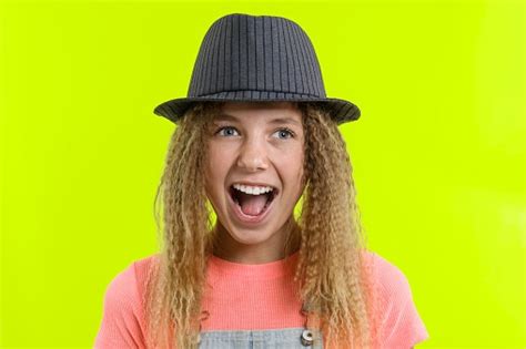 모자와 함께 카메라를 보고 곱슬 머리 놀된 행복 한 십 대 소녀 노란색 스튜디오 배경 위에 입을 열으십시오 곱슬 머리에 대한