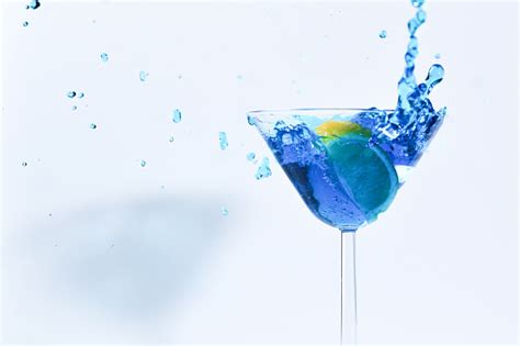 유리에 푸른 액체와 칵테일 물튀김과 방울로 액체를 붓는 푸른 물이있는 유리 흰색 배경에 스플래시와 알코올로 채우는 마티니 유리