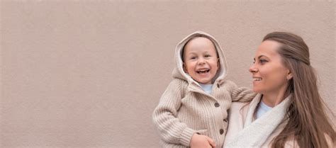 그녀의 팔에 어린 소년 35 세를 들고 젊은 어머니 여자 행복 미소 웃음 휴식과 재미 여유 공간 배경 벽 후드와 따뜻한 의류 베이지 색 의류 스웨터 2명에 대한 스톡 사진