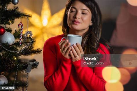 크리스마스 트리 근처에 서서 뜨거운 음료 한 잔을 들고 집에서 휴가 분위기를 즐기는 아름다운 젊은 여자 겨울에 대한 스톡 사진 및 기타 이미지 Istock