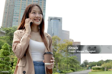 양복을 입은 행복 한 아시아 사업가가 휴대 전화와 종이 커피 컵을 홀링하고 푸른 정원에서 일하고 고층 빌딩 사무실을 흐릿한 배경으로 사용합니다 공립공원에서 스마트폰을 이용해