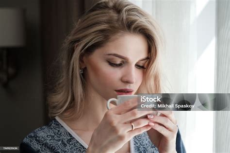 창에 의해 파란색 가운에 아름 다운 젊은 금발 여자 접시와 함께 화이트 컵에서 커피 또는 차를 마신다 아침 햇살 침실 창 가정 생활에 대한 스톡 사진 및 기타 이미지