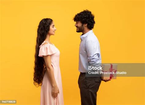 흥분한 인도 남자는 포장된 선물 상자를 들고 있고 등 뒤에 여자 친구를 위한 선물을 숨기고 있다 노란색 배경 20 24세에 대한 스톡 사진 및 기타 이미지 Istock