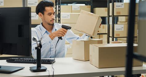 행복한 아시아 남자는 공식적인 유니폼 셔츠를 입고 책상 앞에 앉아 컴퓨터와 바코드 기계 스캐너를 사용하여 창고에서 골판지 상자 배달에 고객 온라인 데이터를 기록합니다