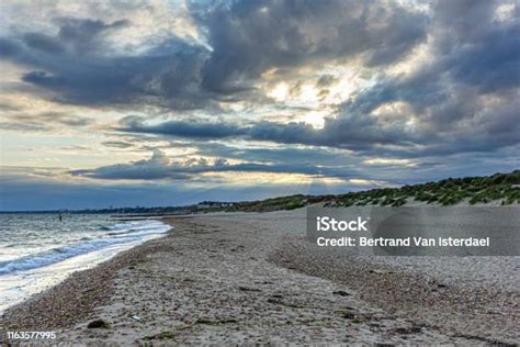 모래 해변을 따라 인상적인 어둡고 밝은 구름을 가로 지르는 태양과 빛의 광선 고르지 않은 푸른 물 녹색 식물이있는 모래 언덕 및 배경에있는 도시와 함께 폭풍우가 몰아치는