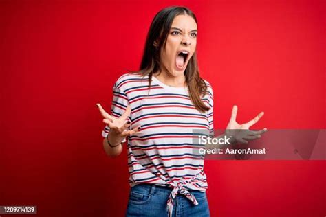 젊은 아름 다운 갈색 머리 여자 캐주얼 줄무늬 티셔츠 를 입고 빨간 배경 미친 미친 위에 서 미친 소리와 공격적인 표정과 팔을