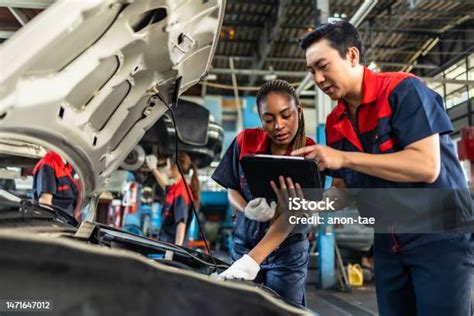 차고의 리프터에서 차량 상태를 확인하는 엔지니어 팀 제복을 입은 젊은 자동차 정비사가 카메라를 바라보며 웃으며 차를 살펴보고 있다 20 24세에 대한 스톡 사진 및 기타