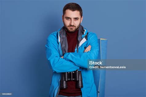 접힌 손으로 파란색 배경 위에 고립 된 수염을 가진 잘 생긴 남자의 수평 샷 매트와 쌍안경을 가진 여행자 재킷을 입고 카메라를보고 분노와 침을 표현한다 관광객에 대한 스톡