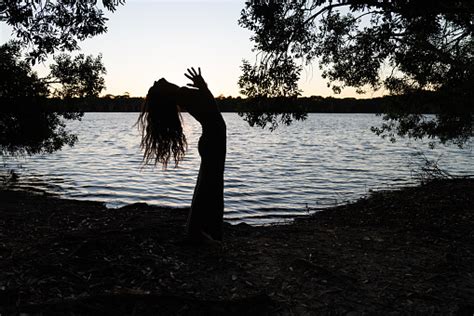 요가 포즈를 취하는 여성의 전체 길이 실루엣 샷 그녀는 다시 구부러지고 그녀의 머리카락이 뒤에서 흐르고 배경에는 나무와 호수가 있습니다 40 44세에 대한 스톡 사진 및
